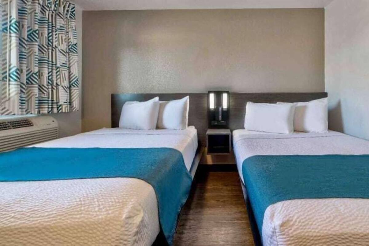 Quarto de hotel com duas camas, uma mesa de cabeceira, televisão presa à parede e janela com cortinas.