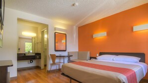 Quarto de hotel com cama, escrivaninha, cadeira e banheiro visível por uma porta aberta.