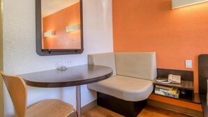 Quarto pequeno e moderno com uma mesa redonda, um banco estofado, um espelho e uma estante com um telefone e livros.