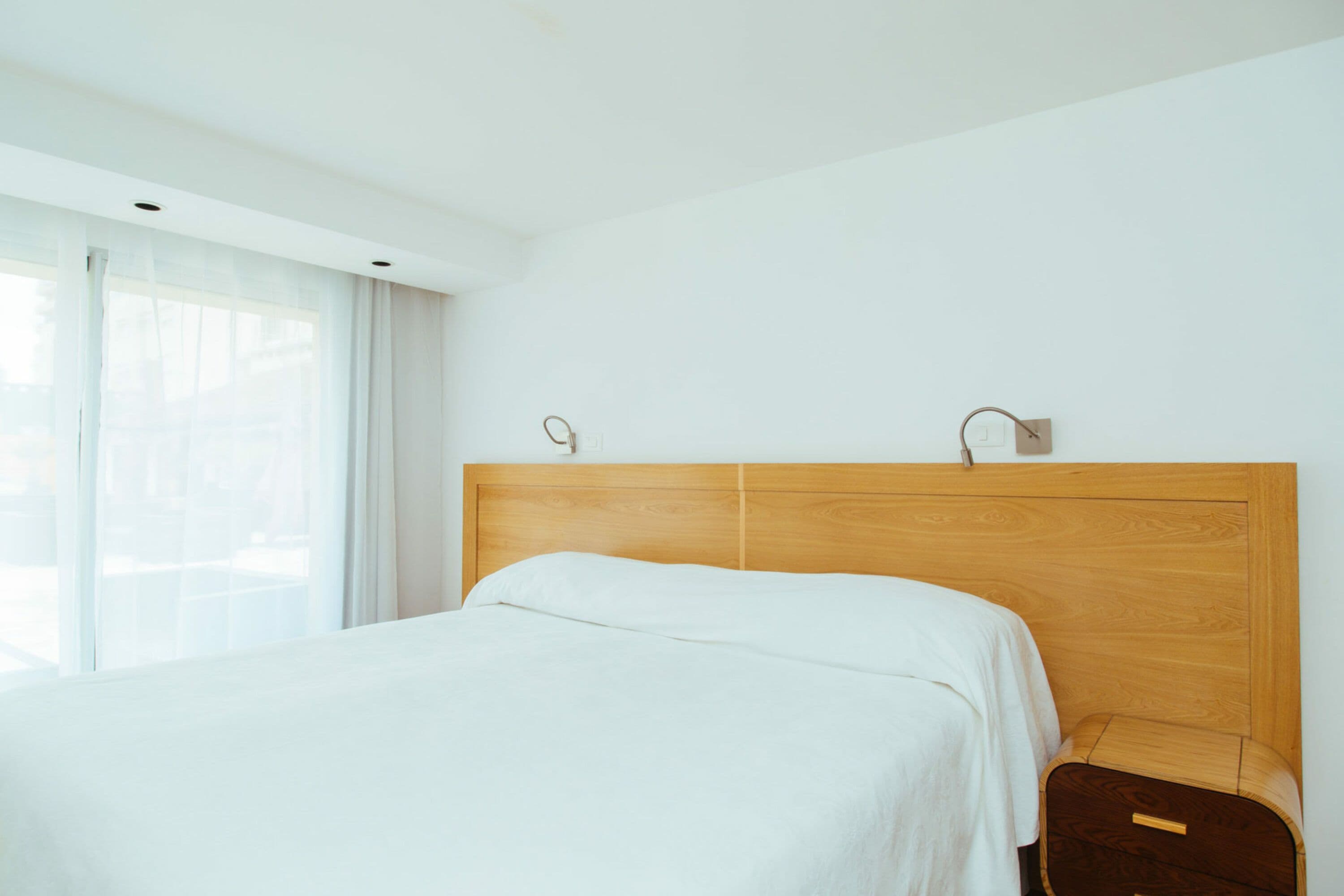 Quarto de hotel com cabeceira de madeira, cama com roupa de cama branca, mesa de cabeceira e janela com cortinas finas.