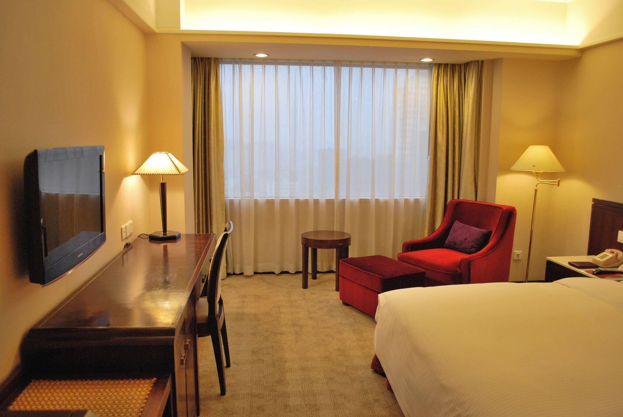 Quarto de hotel com uma cama, uma escrivaninha com cadeira, uma poltrona vermelha, uma mesa pequena, uma televisão na parede e uma janela com cortinas.