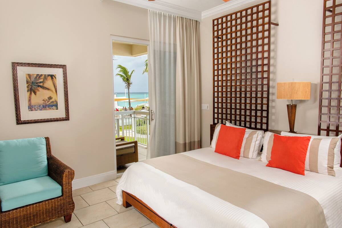 Quarto de hotel com uma cama, uma cadeira de vime, vista para a praia e um painel de madeira.