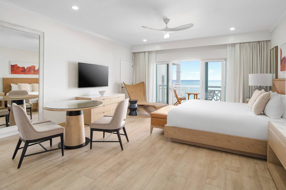 Quarto de hotel com cama, televisão, mesa de jantar com cadeiras, varanda com vista para o mar e ventoinha no teto.