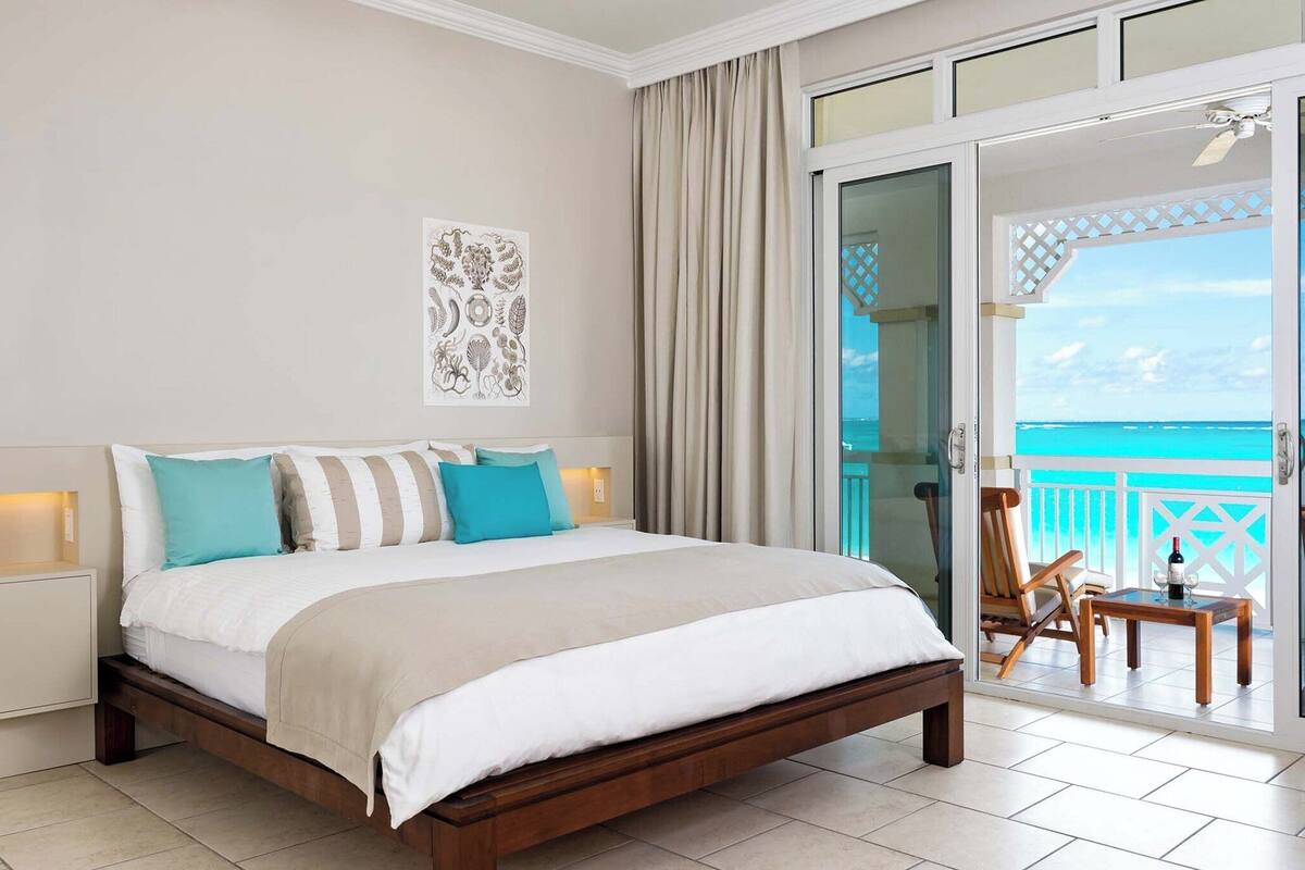 Quarto com cama, mesa de cabeceira, janela com cortinas e vista para o mar.