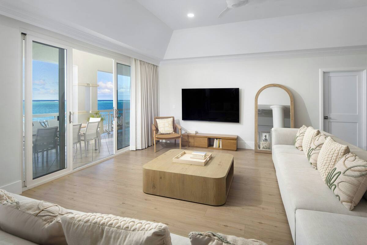 Uma sala de estar ampla com uma grande porta de vidro deslizante que dá para uma varanda, uma TV de tela plana fixada na parede e uma mesa de centro de madeira.