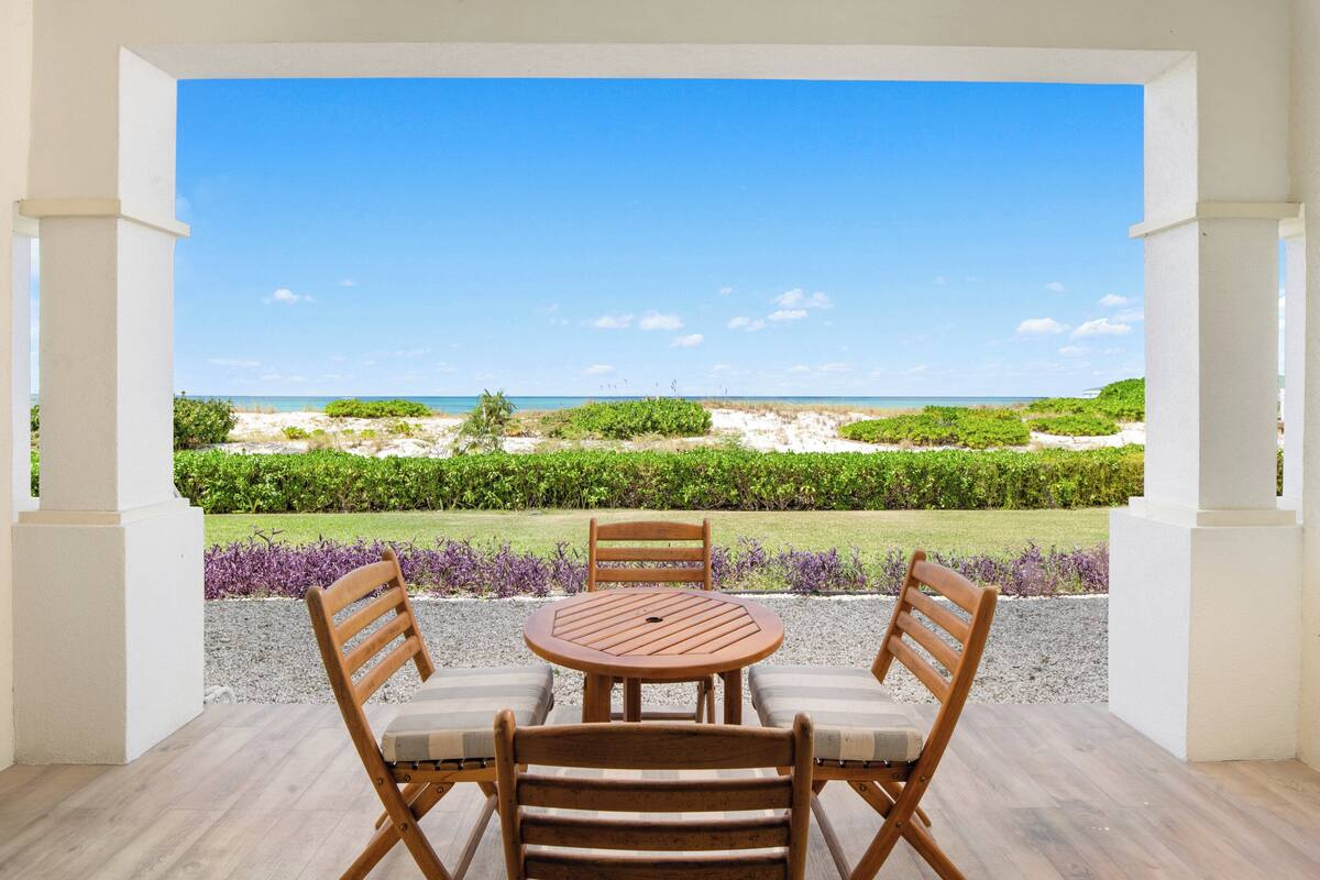 Uma área externa com mesas e cadeiras, com vista para a praia e vegetação.