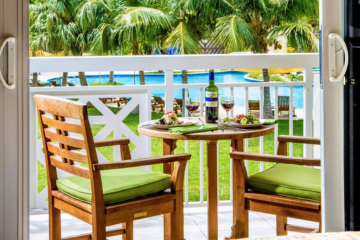 Uma cadeira de madeira com almofada verde, uma mesa de madeira com uma garrafa e copos, e vista para uma piscina e palmeiras.