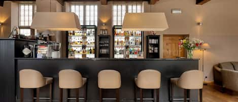 Uma área de bar com quatro banquetas, piso de madeira e um balcão abastecido com diversas garrafas.