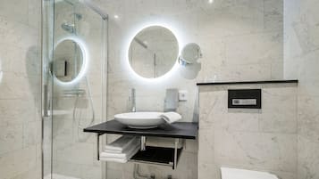 Uma casa de banho moderna com cabine de duche de vidro, espelho redondo, lavatório branco e bancada preta.