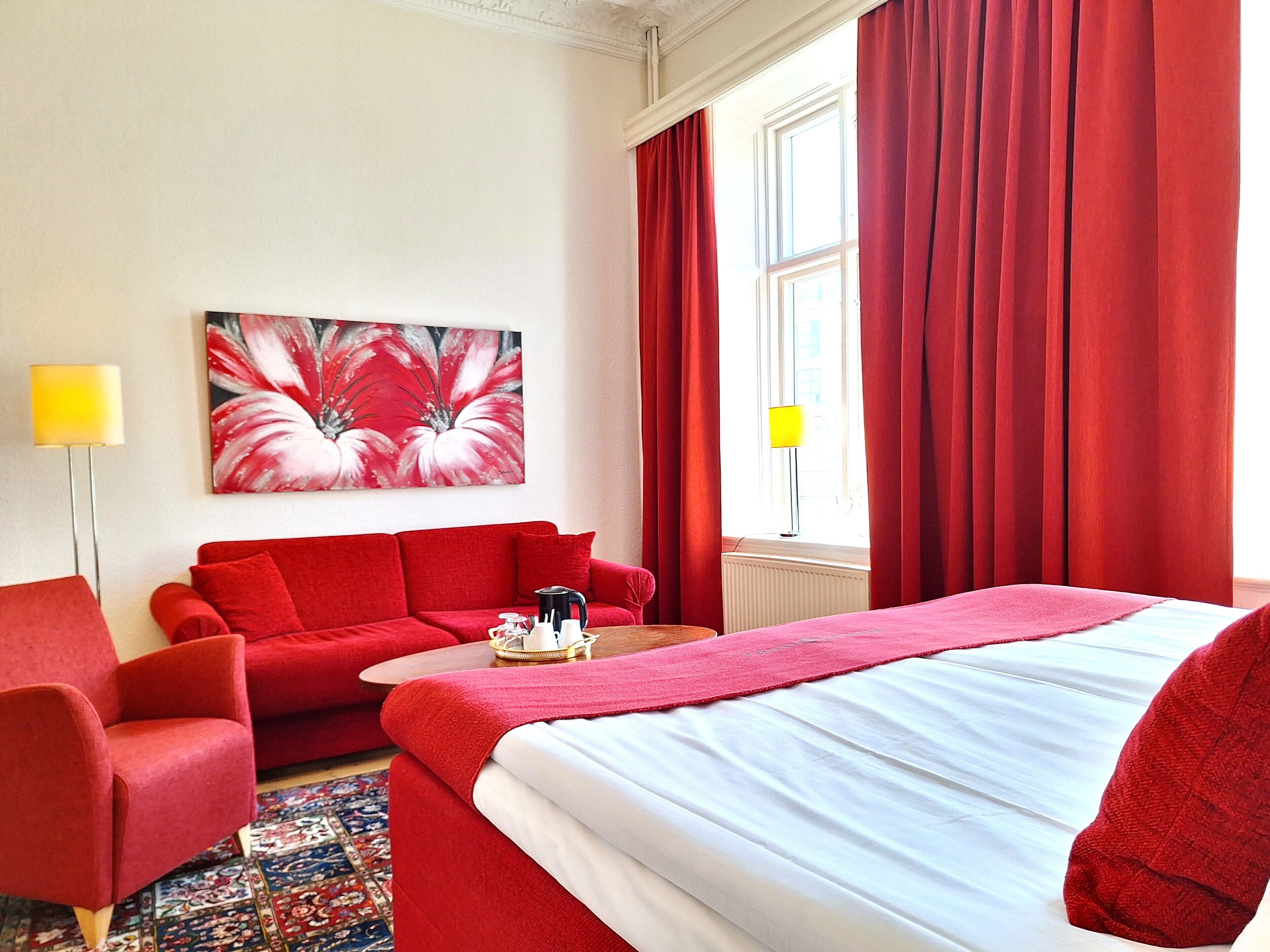 Quarto de hotel com uma cama grande, um sofá vermelho, uma mesa de centro e um quadro na parede.