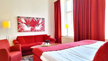 Quarto de hotel com uma cama grande, um sofá vermelho, uma mesa de centro e um quadro na parede.