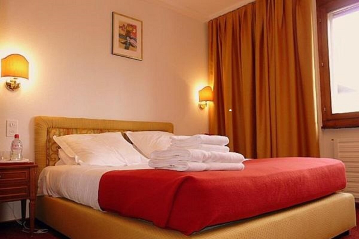 Quarto de hotel com cama, mesa de cabeceira, luminária, janela com cortinas e um quadro na parede.