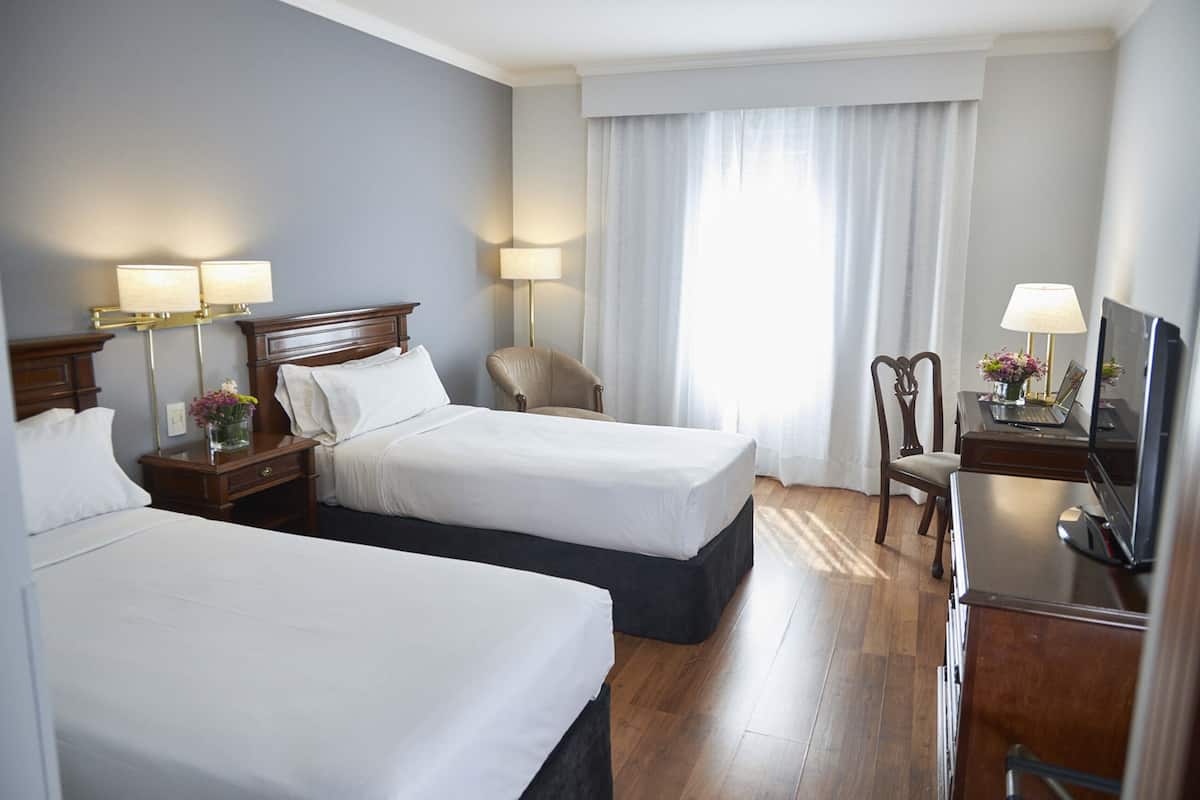 Quarto de hotel com duas camas, uma mesa de trabalho com televisão, uma cadeira e duas luminárias de cabeceira.