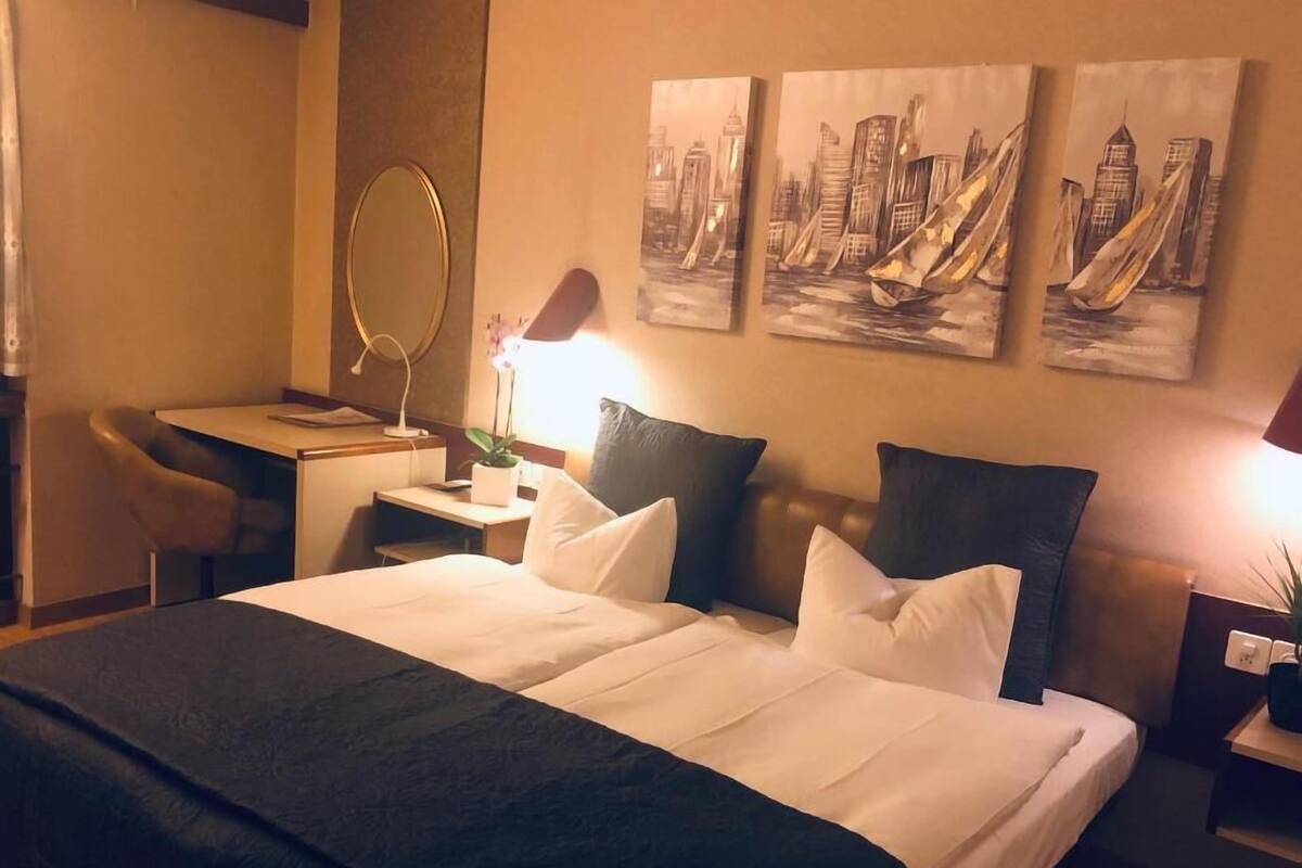 Quarto de hotel com cama, escrivaninha, cadeira, espelho e três quadros de veleiros na parede.