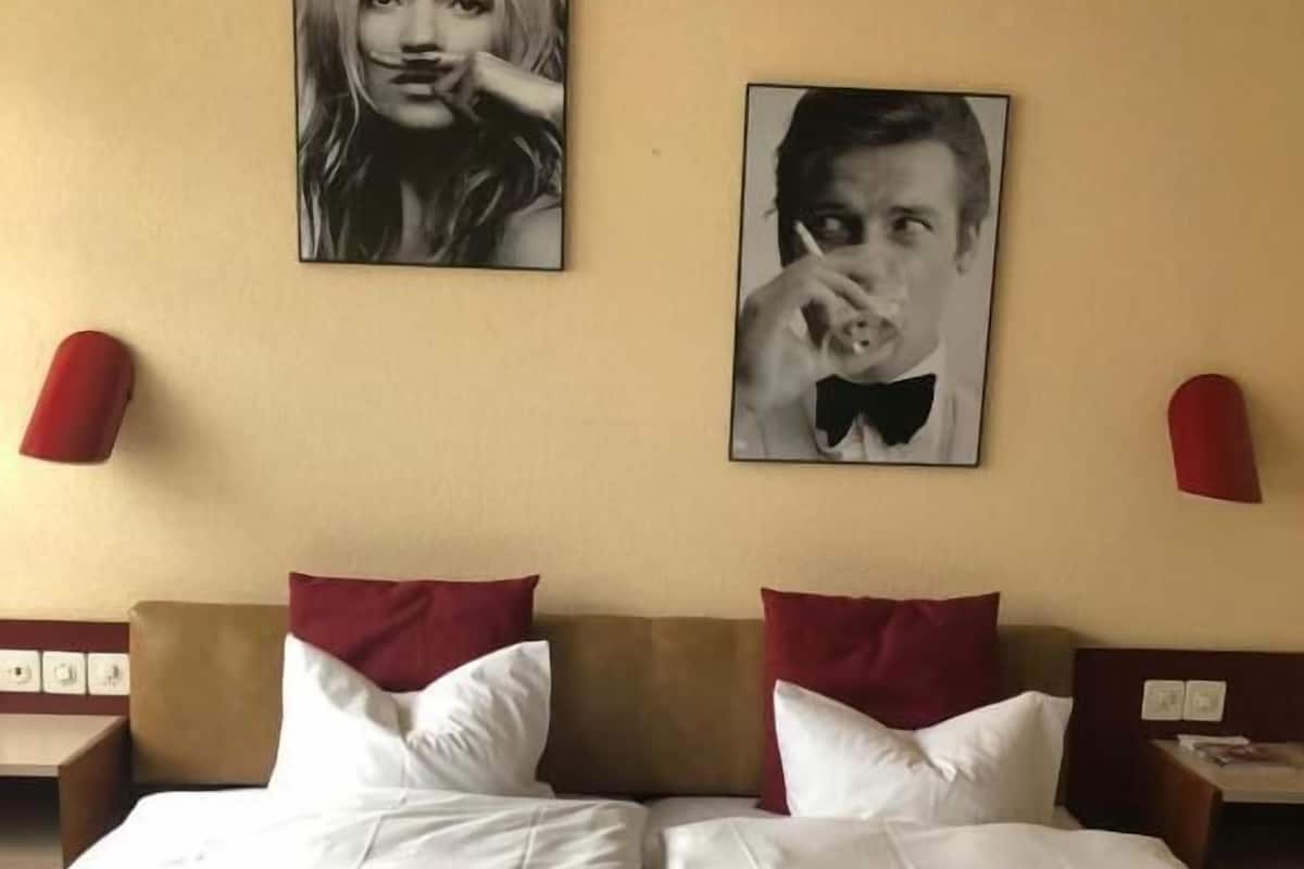 Quarto de hotel com cama grande, roupa de cama vermelha e branca, duas luminárias de parede vermelhas e duas imagens emolduradas na parede.