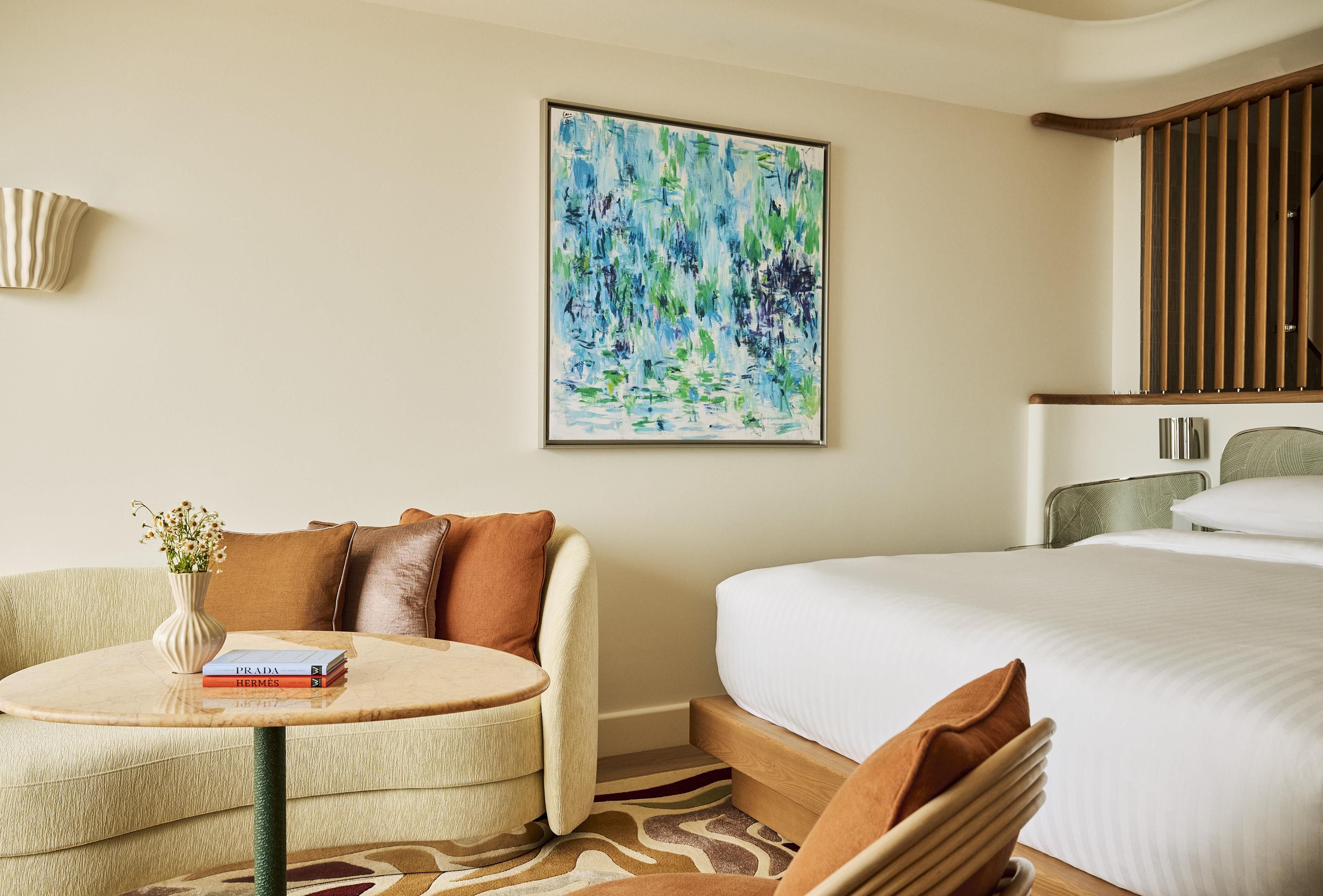 Quarto de hotel com uma cama, uma mesa redonda com um vaso e livros, um quadro na parede e uma escadaria.