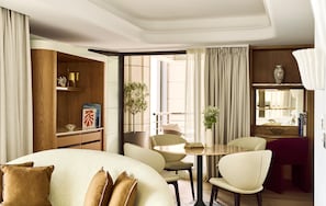 Quarto de hotel com cama, mesa de jantar com cadeiras, estante de livros e uma janela grande com cortinas.
