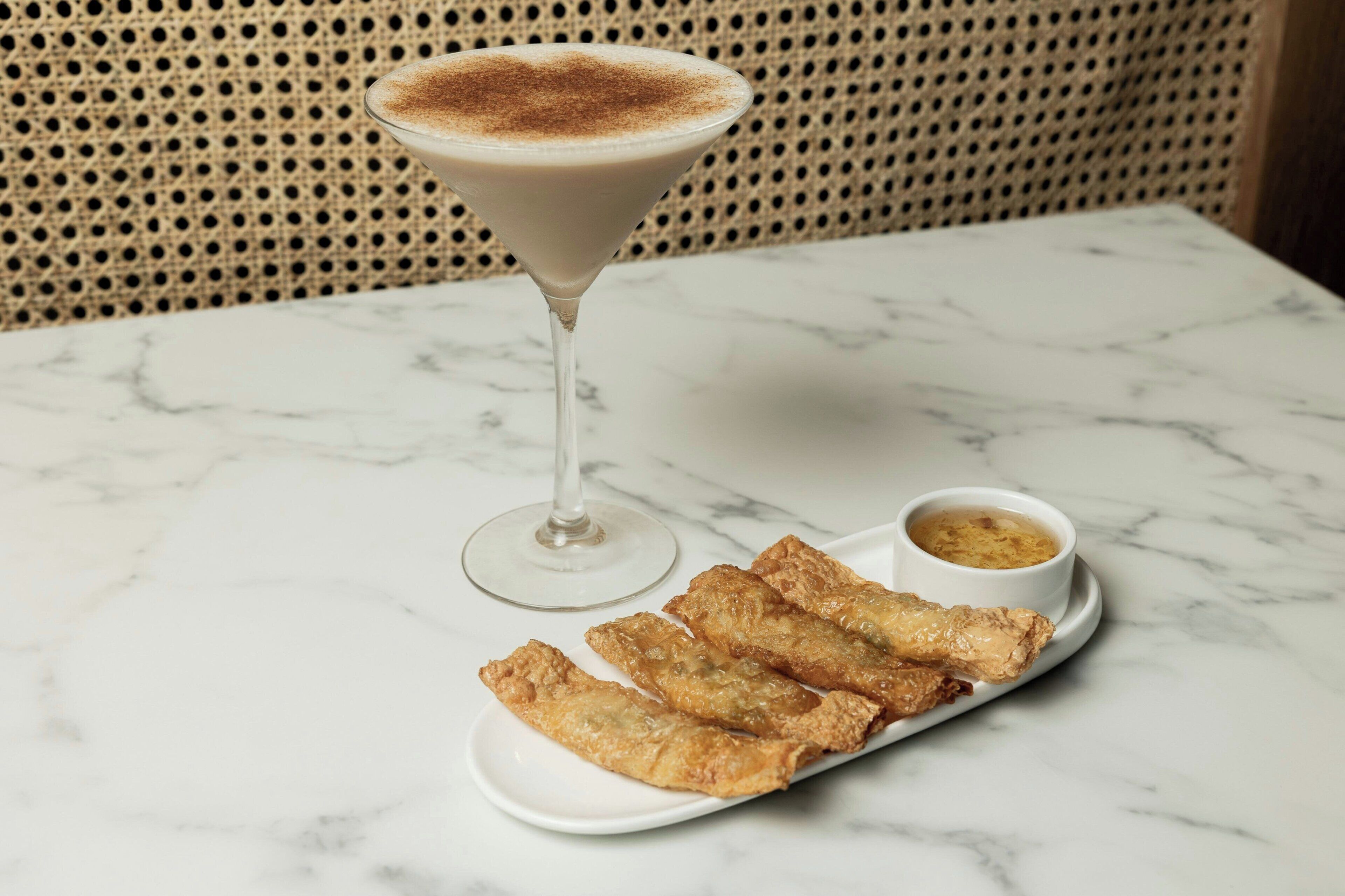 Um copo de martini com espuma, um prato de doces fritos e uma pequena tigela de molho sobre uma mesa de mármore.