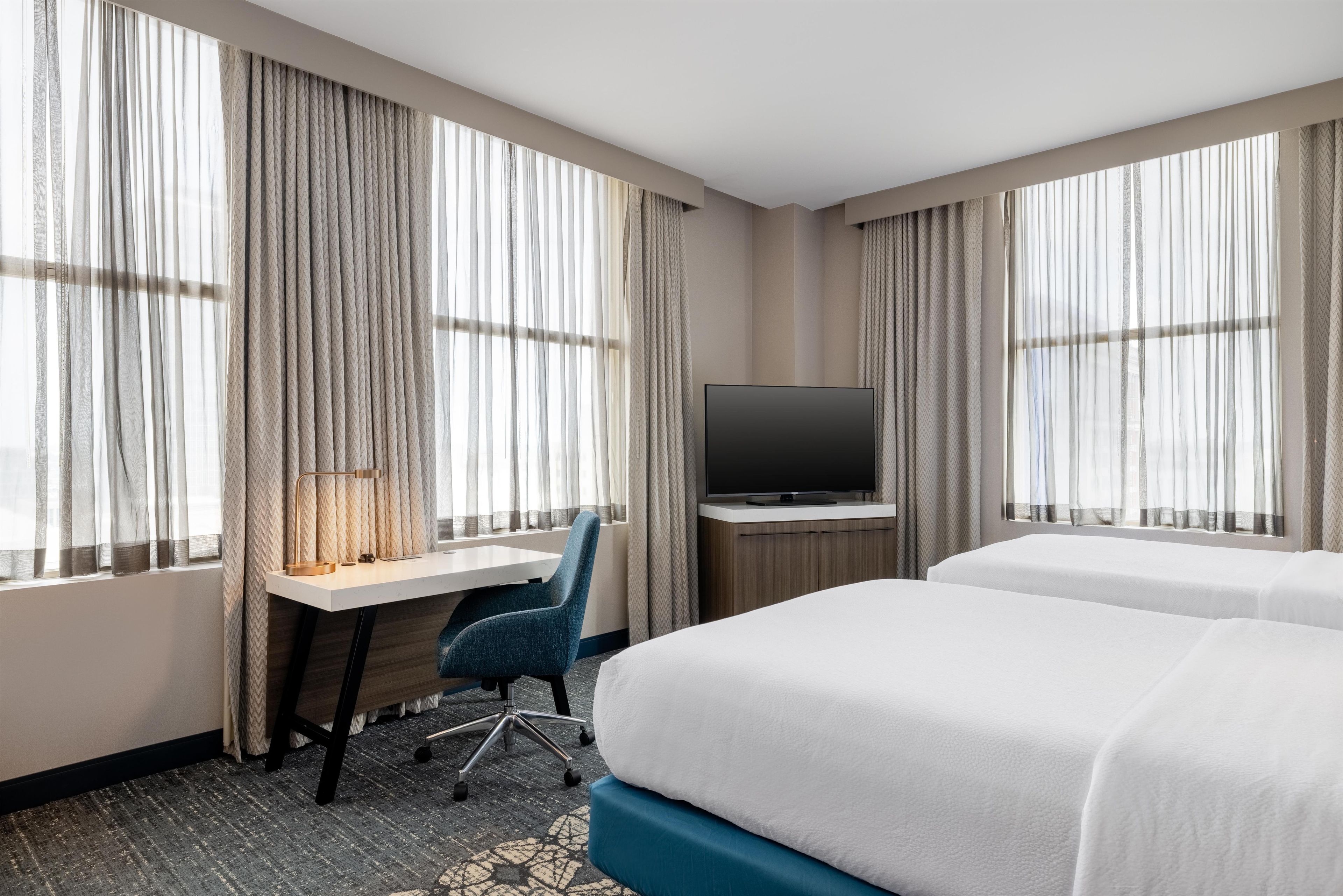 Quarto de hotel com cama, escrivaninha, cadeira, televisão e cortinas.