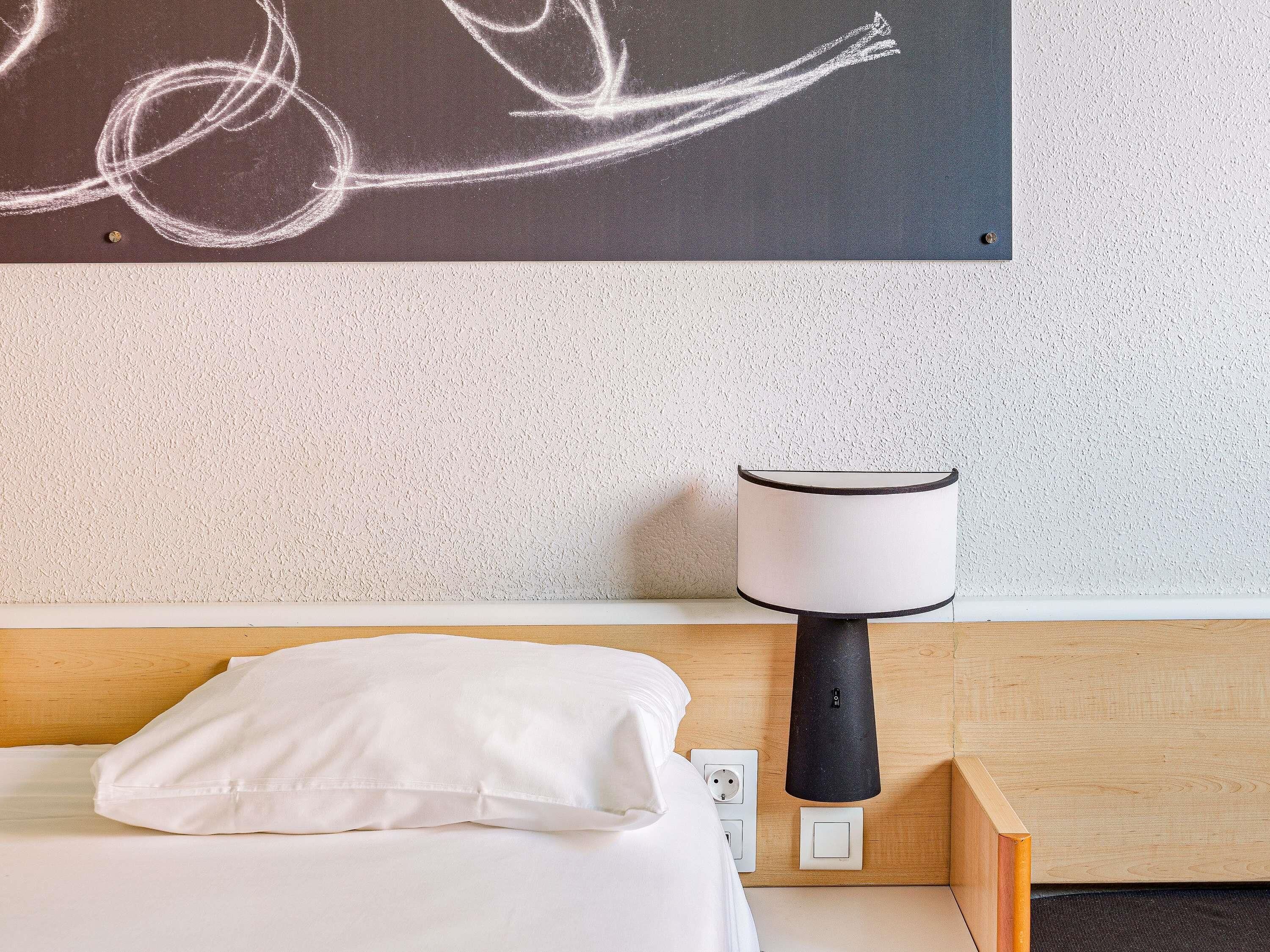Quarto de hotel com uma cama, um travesseiro branco, uma luminária fixada na parede e um quadro negro com desenhos abstratos.