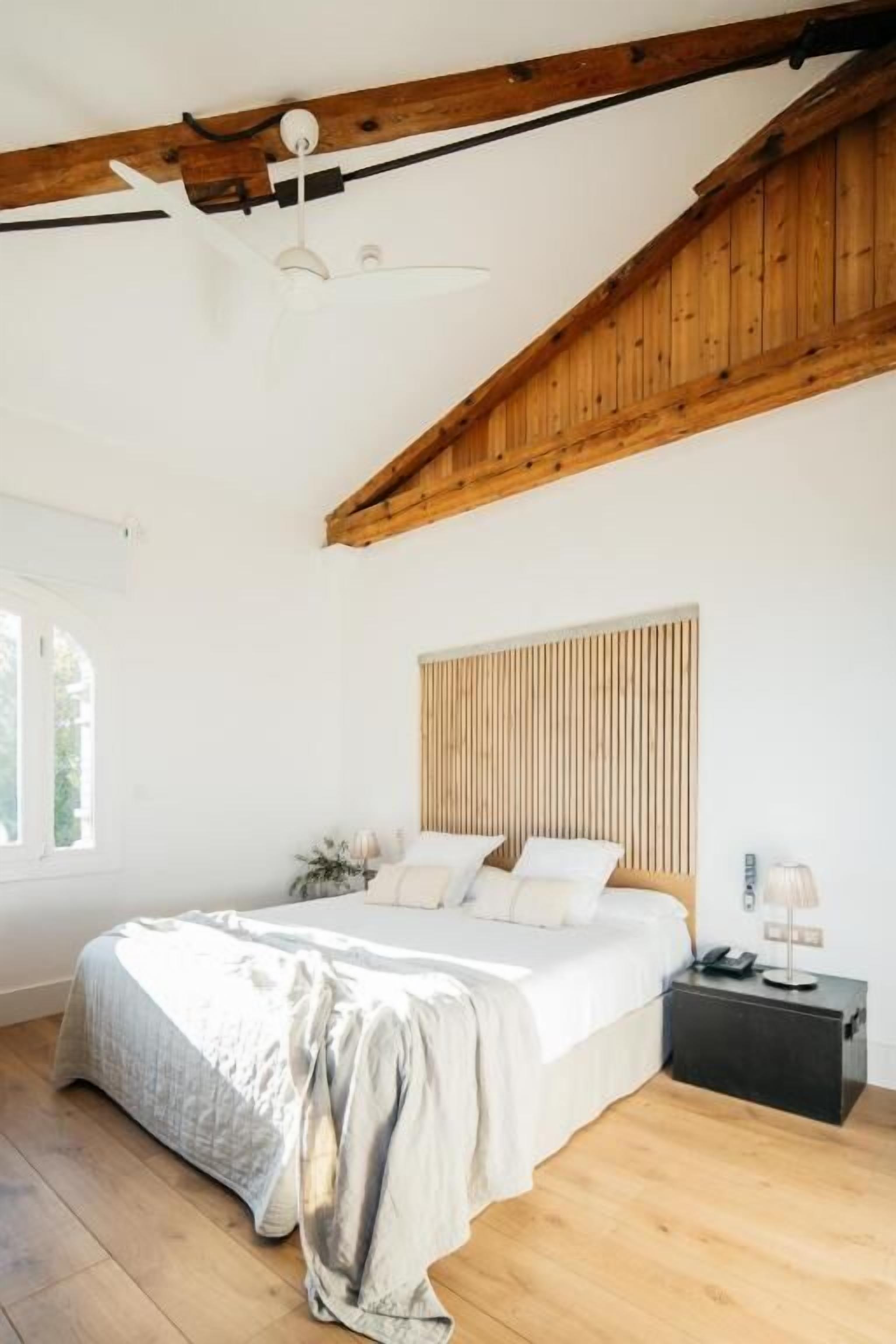 Quarto com teto de madeira, cama com roupa de cama branca, mesa de cabeceira com abajur e janela que proporciona luz natural.