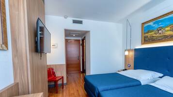 Quarto de hotel com piso de madeira, duas camas com roupa de cama azul, uma cadeira vermelha, televisão fixada na parede e um quadro sobre a cama.