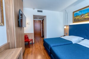 Quarto de hotel com piso de madeira, duas camas com roupa de cama azul, uma cadeira vermelha, televisão fixada na parede e um quadro sobre a cama.