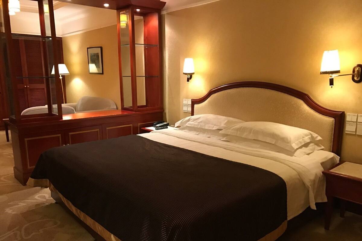 Quarto de hotel com uma cama grande, cabeceira de madeira, mesa de cabeceira, escrivaninha com cadeira e espelho.