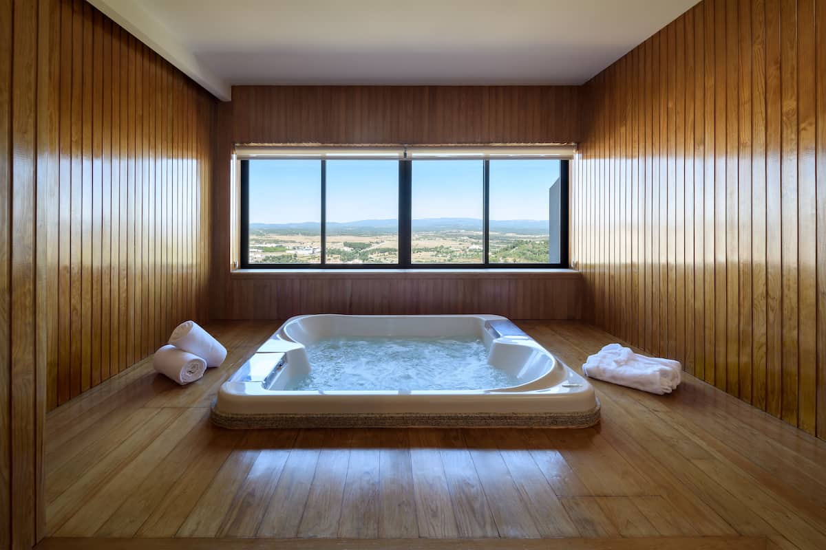 Quarto com painéis de madeira, uma janela grande, hidromassagem e toalhas no chão.
