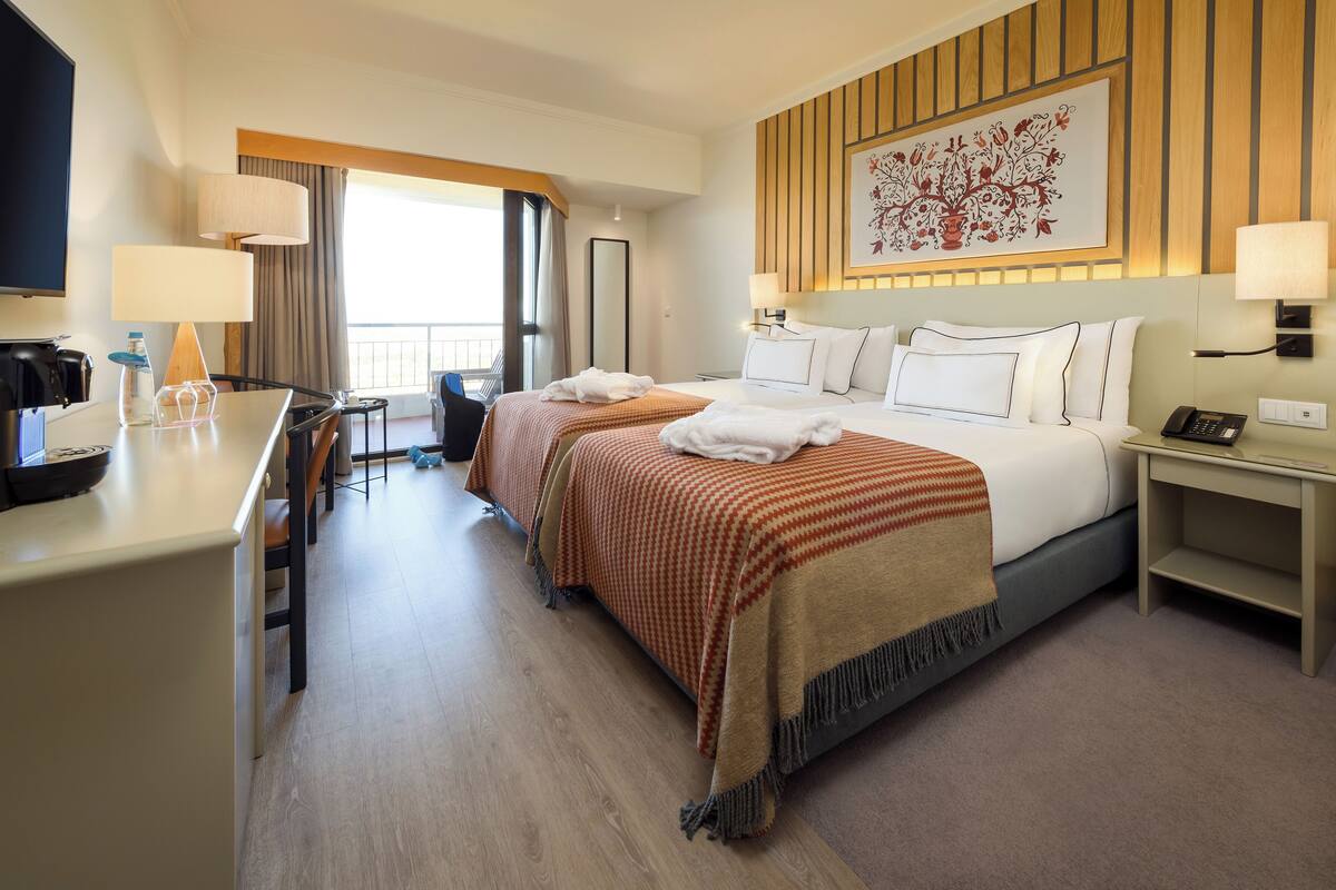 Quarto de hotel com cama, mesas de cabeceira, escrivaninha, cadeira, televisão e janela com cortinas.