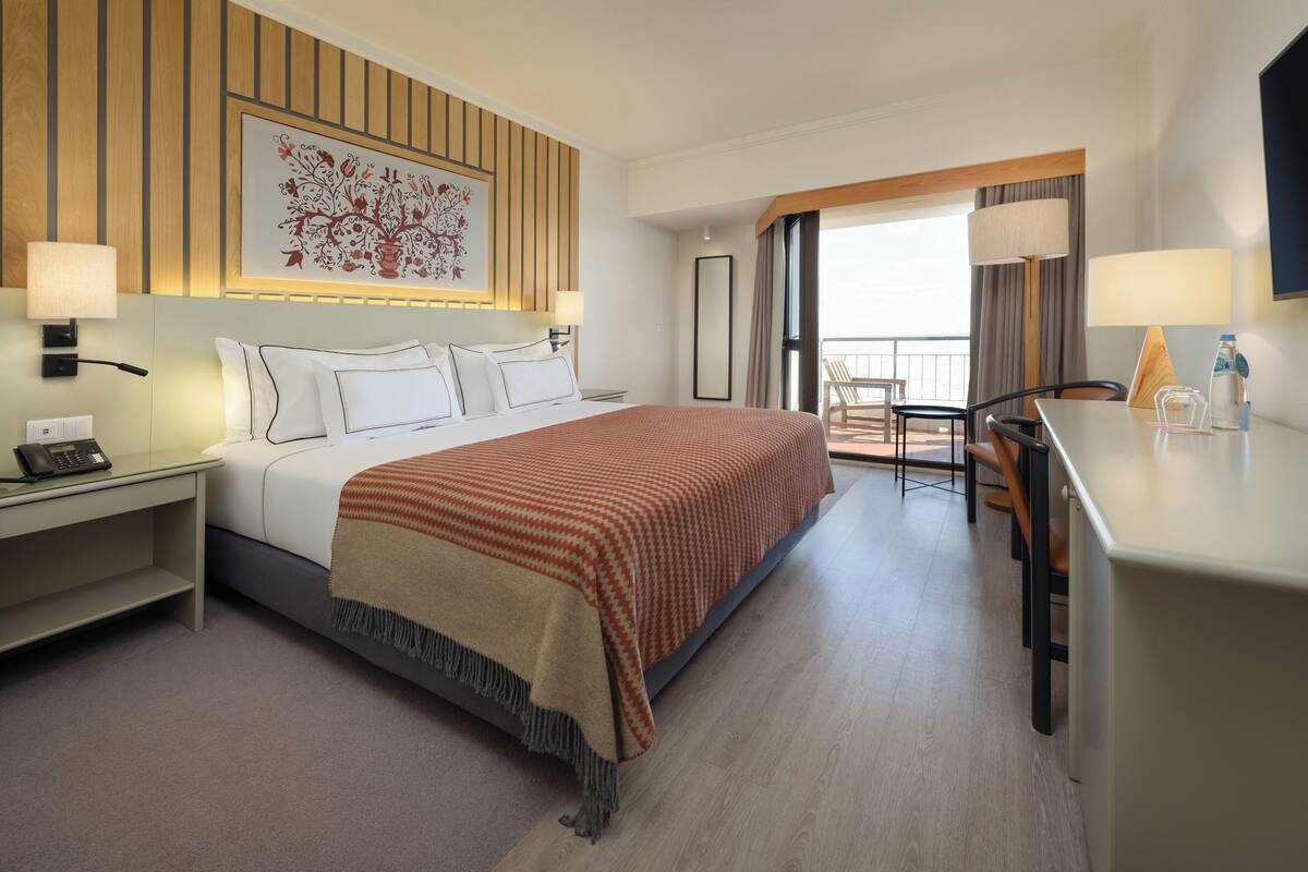 Quarto de hotel com cama, mesas de cabeceira, escrivaninha, cadeira, varanda e uma janela grande.