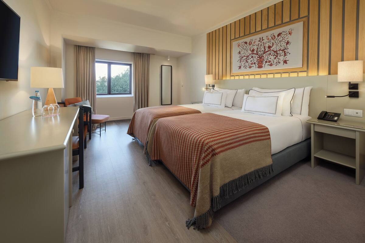 Quarto de hotel com duas camas, uma escrivaninha, uma cadeira, uma janela grande e uma obra de arte na parede.