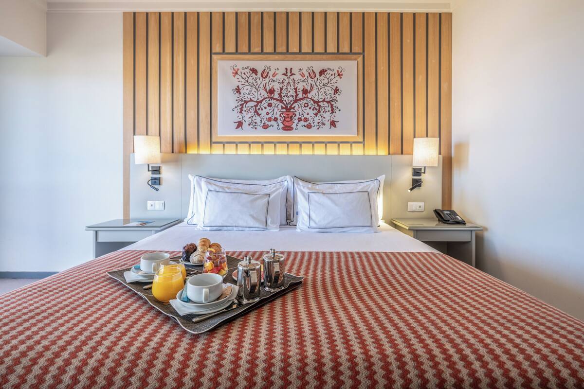 Quarto de hotel com uma cama, duas mesas de cabeceira, um suporte com itens de café da manhã e uma obra de arte pendurada na parede.