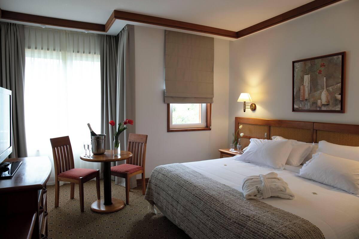 Quarto de hotel com uma cama, uma escrivaninha com cadeira, uma mesinha com um vaso e flores, um abajur e um quadro na parede.