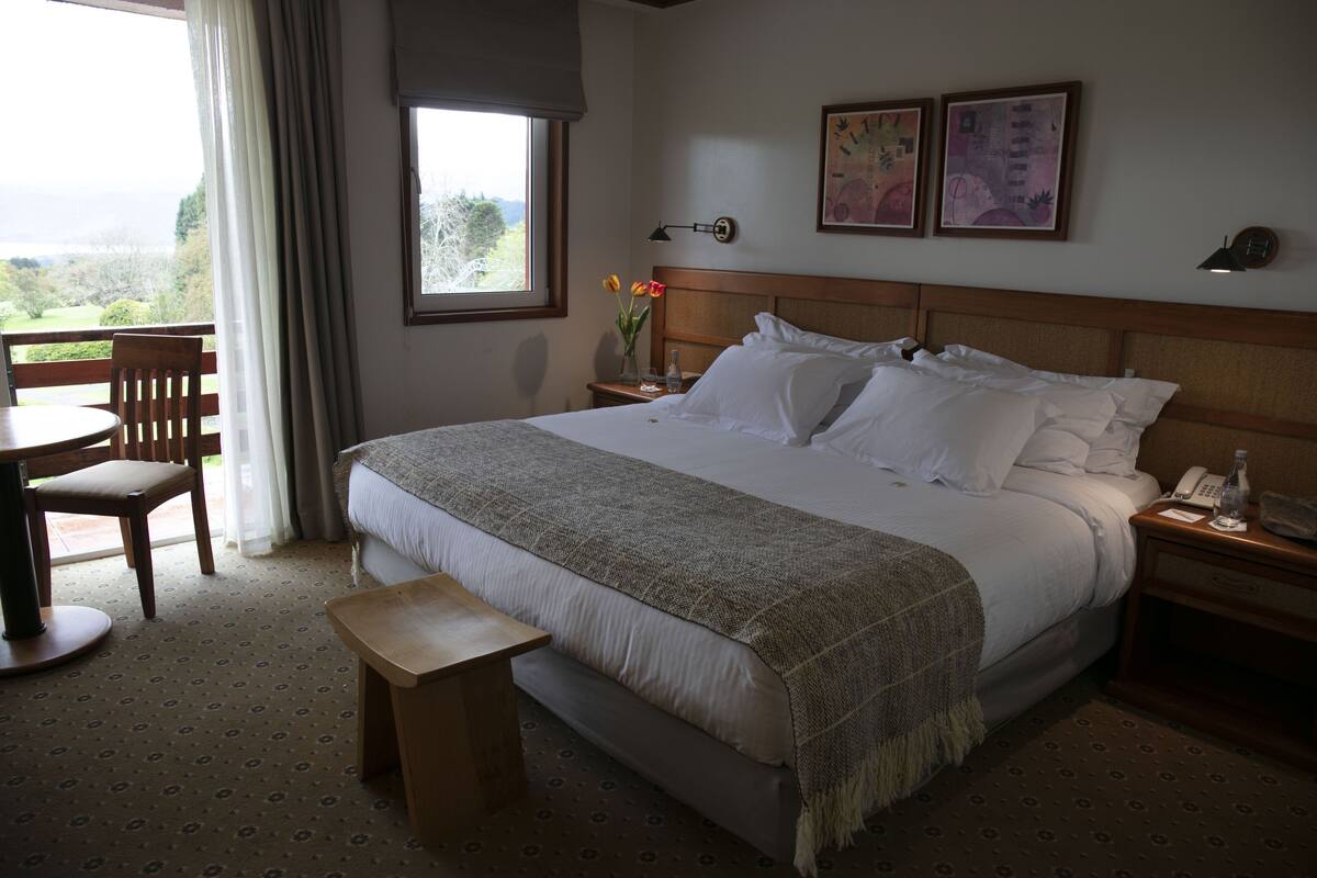 Quarto de hotel com uma cama grande, uma mesa de cabeceira, uma mesinha, uma cadeira e uma janela com vista para o exterior.