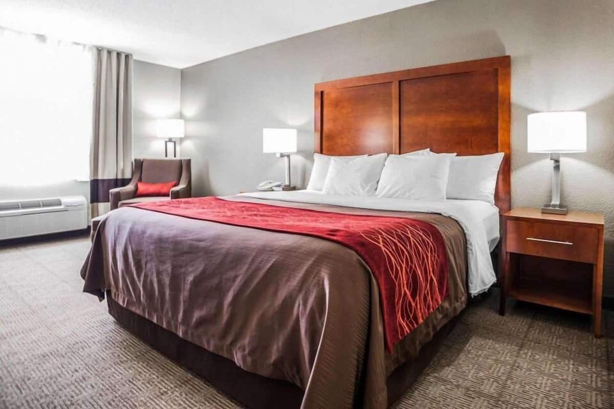 Quarto de hotel com uma cama grande, duas mesas de cabeceira com abajures, uma cadeira e uma janela com cortinas.