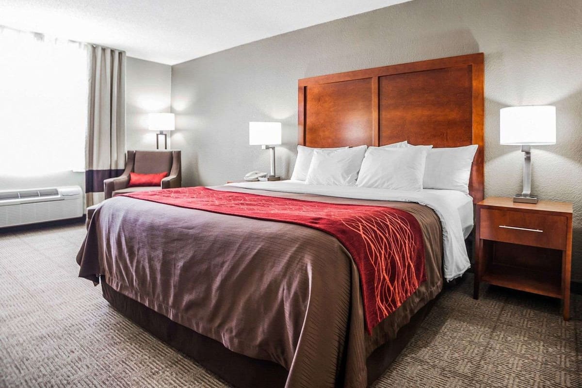 Quarto de hotel com uma cama grande, duas mesas de cabeceira com abajures, uma cadeira e uma janela com cortinas.