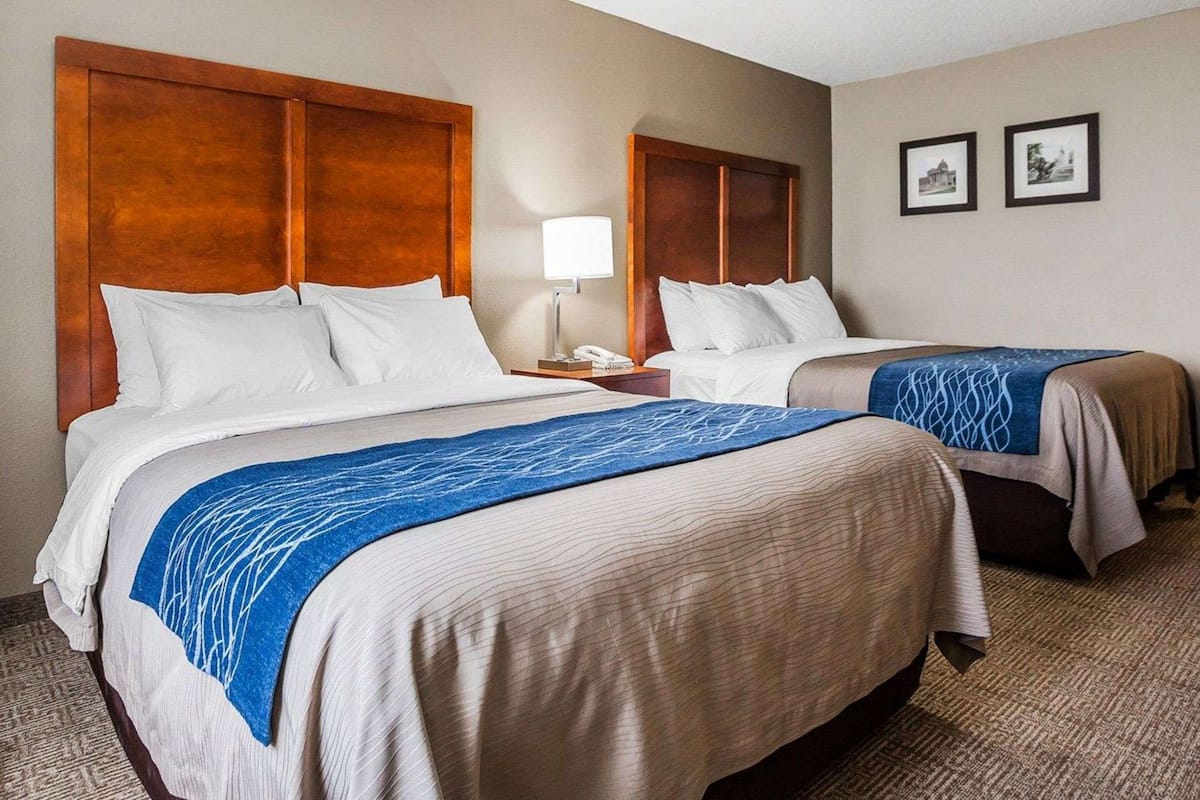 Quarto de hotel com duas camas, cabeceiras de madeira, roupa de cama branca, uma mesa de cabeceira com abajur e duas imagens emolduradas na parede.