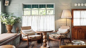 Um quarto aconchegante com piso de madeira, uma mesa redonda, duas cadeiras, uma mesinha de vime, uma planta em vaso e uma janela com cortinas.