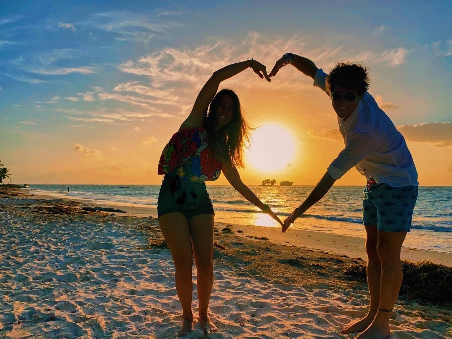 Duas pessoas formando um coração com as mãos na praia ao pôr do sol.