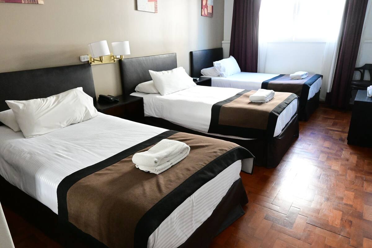 Quarto de hotel com duas camas, cada uma com cabeceira marrom, roupa de cama branca e uma toalha dobrada na cama. Há uma mesa de cabeceira com uma luminária entre as camas e uma janela com cortinas à direita.
