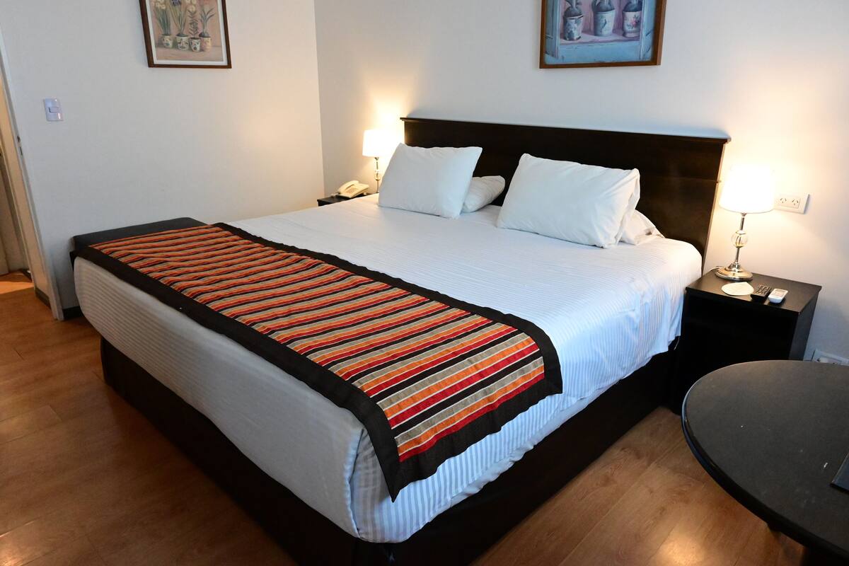 Cama bem arrumada com manta listrada, mesa de cabeceira com abajur e uma pequena mesa redonda.
