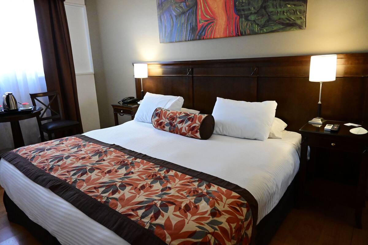 Quarto de hotel com uma cama grande, duas mesas de cabeceira, uma mesa de apoio, uma escrivaninha com cadeira e um quadro na parede.