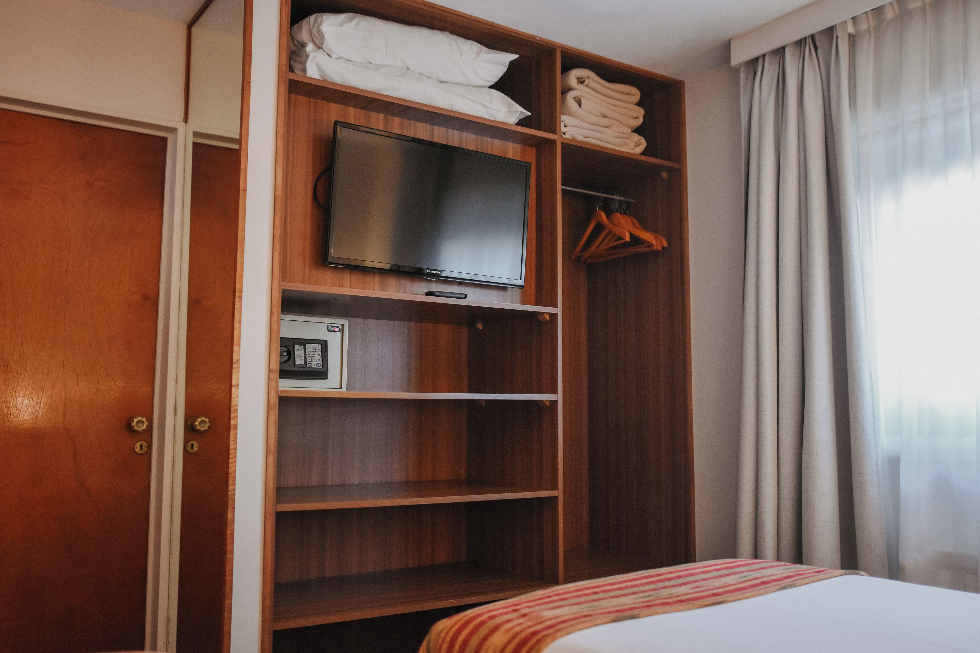 Quarto de hotel com guarda-roupa de madeira, televisão, micro-ondas e cama com colcha listrada.