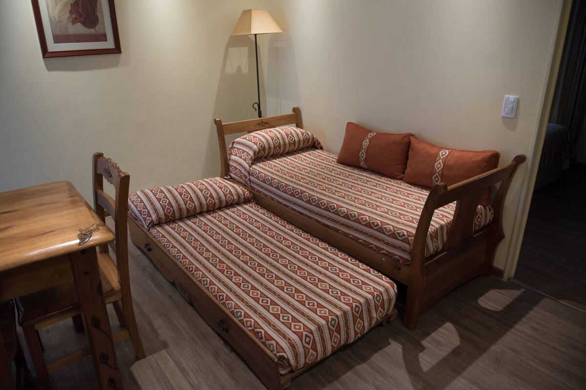 Um quarto pequeno e acolhedor com duas camas de madeira e roupa de cama estampada, uma escrivaninha de madeira e um abajur.