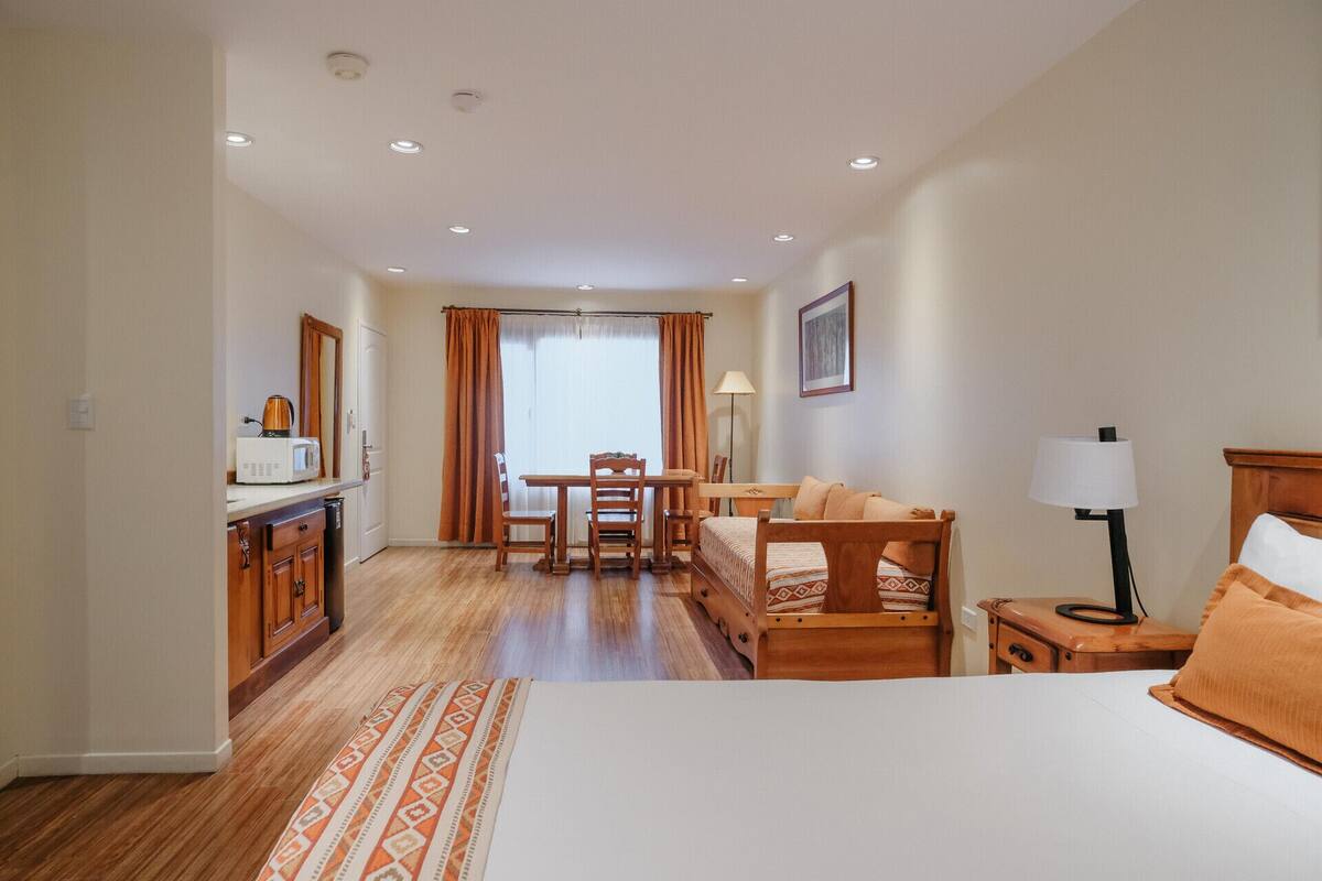 Quarto de hotel com cama, escrivaninha, cadeira, mesa, luminária e cozinha compacta.