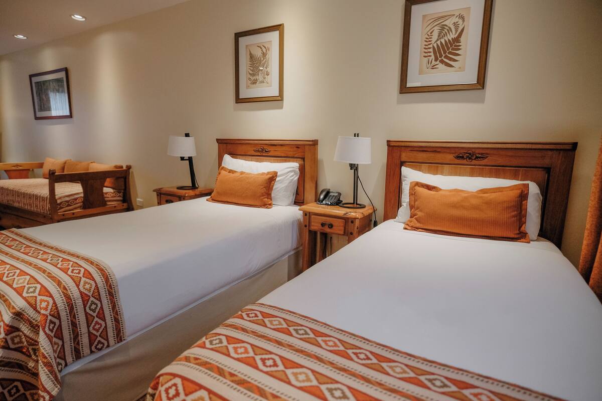 Quarto de hotel com duas camas, cada uma com cabeceira de madeira, roupa de cama branca e colchas com padrão laranja. Há mesas de cabeceira com luminárias e telefone.