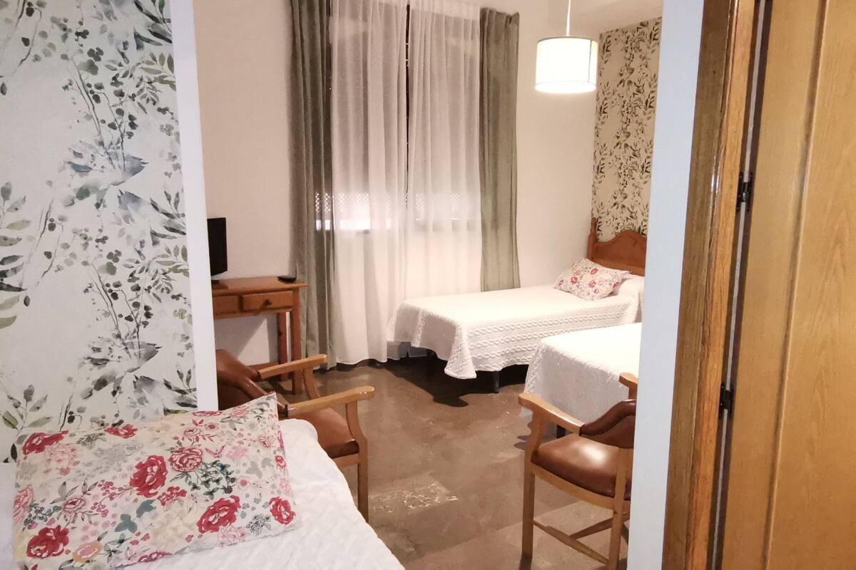 Quarto com duas camas, uma cadeira de madeira, uma mesinha de cabeceira e uma janela com cortinas leves.