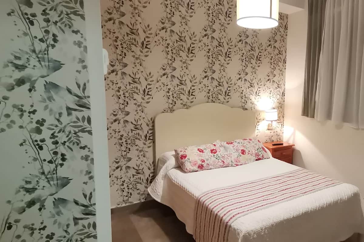 Quarto com cama, mesa de cabeceira, abajur, cadeira e papel de parede floral.