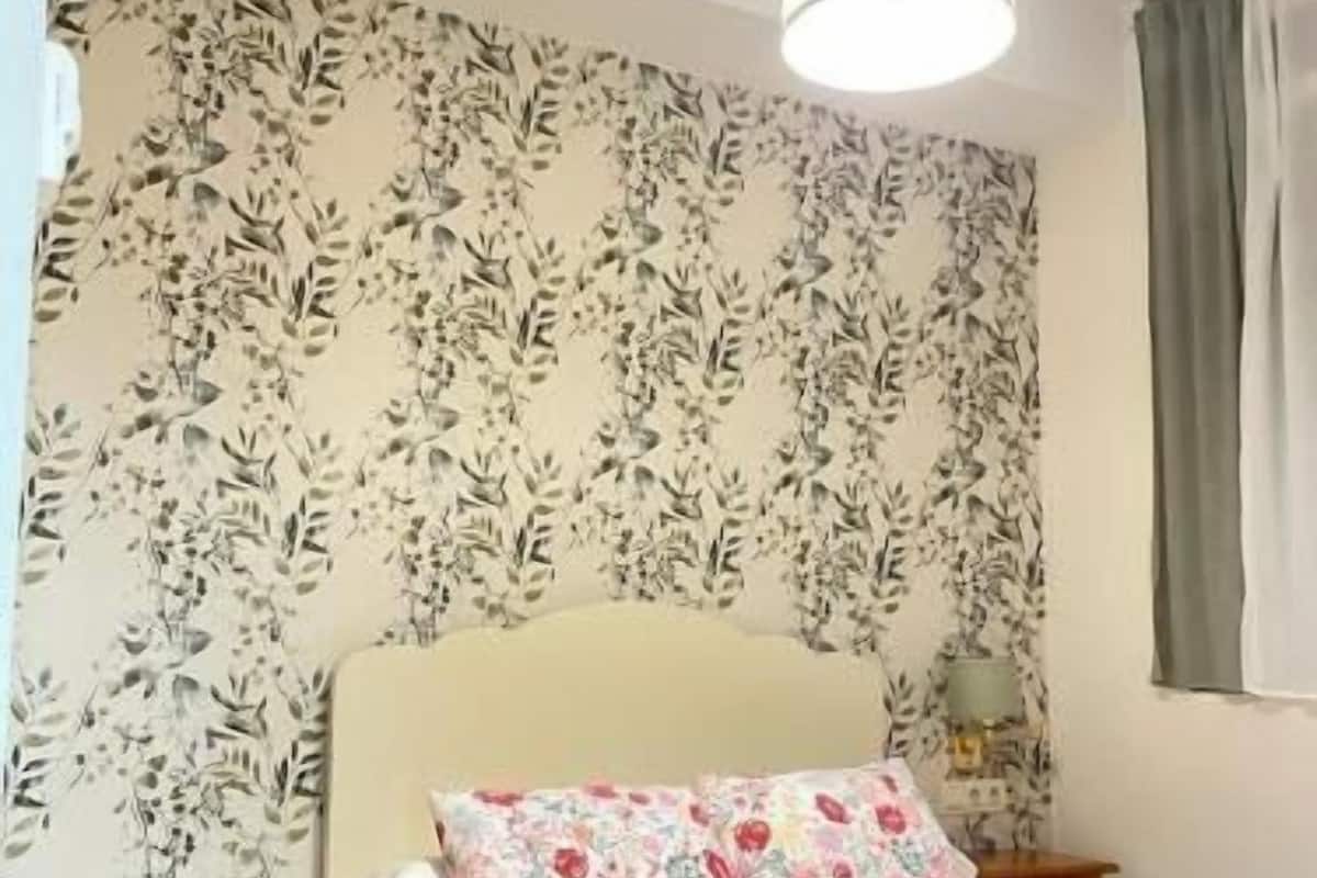 Quarto com cama, papel de parede floral, mesa de cabeceira e luminária pendente.